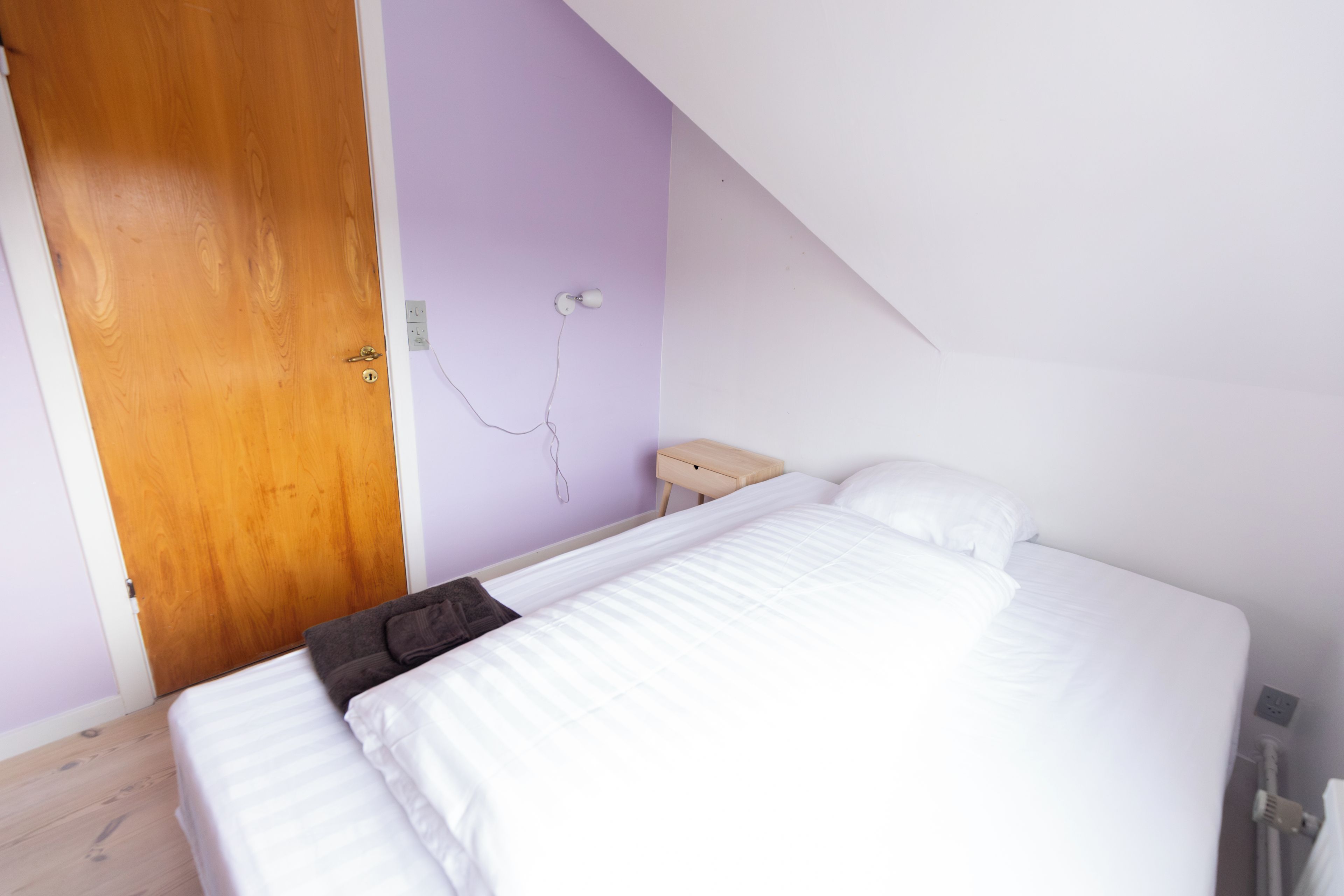 Quarto com porta de madeira, cama branca, mesa de cabeceira e tomada na parede.