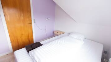 Quarto com porta de madeira, cama branca, mesa de cabeceira e tomada na parede.