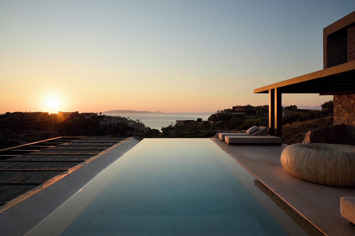 Piscina infinita moderna com vista para o pôr do sol sobre o mar, uma estrutura de pedra e área de estar ao ar livre.