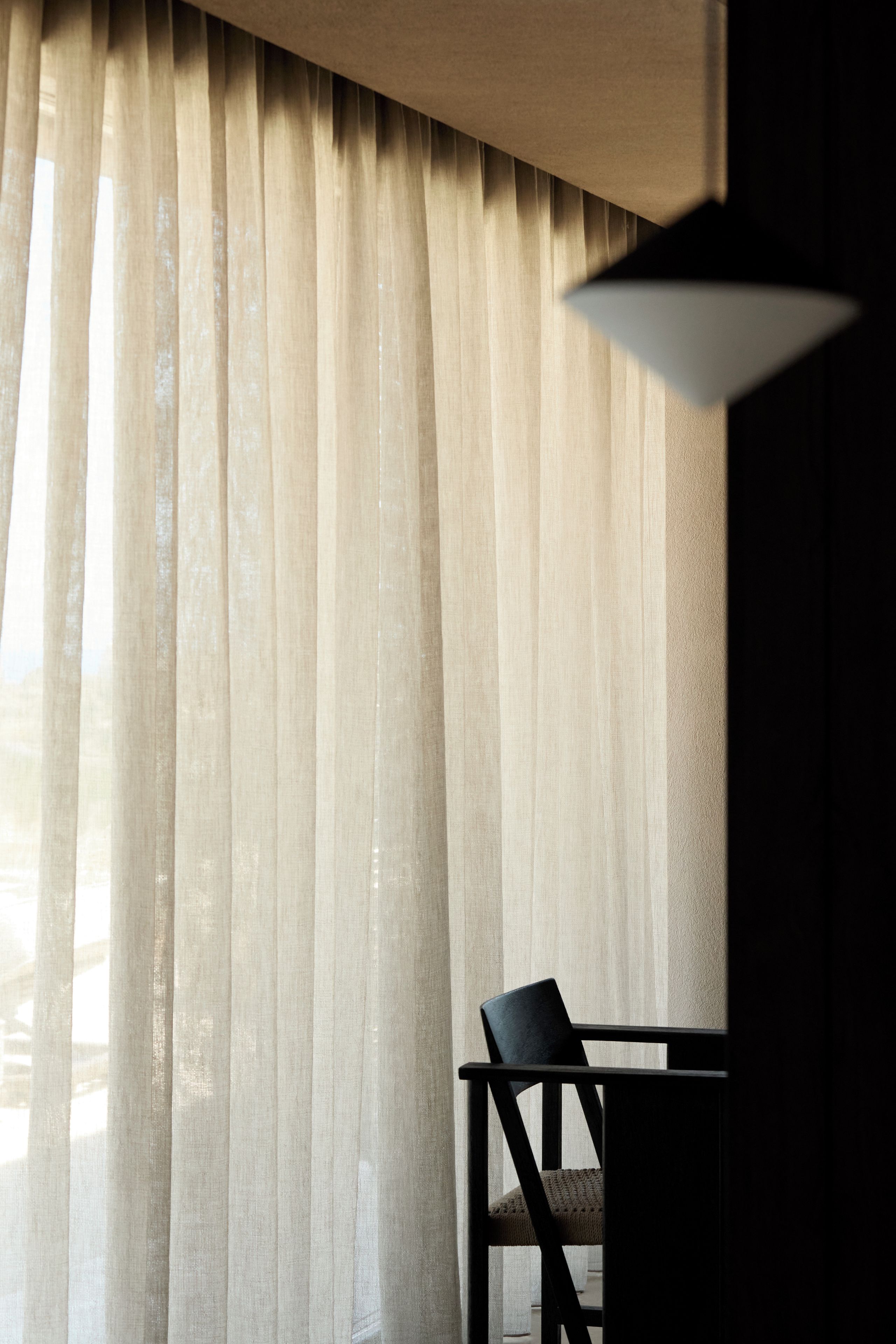 Quarto com cadeira e janela com cortinas leves.