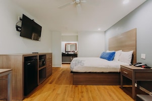 Quarto de hotel com cama, mesa de cabeceira, escrivaninha, televisão na parede e área de cozinha compacta.