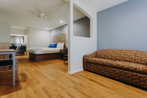 Quarto de hotel moderno com piso de madeira, cama com roupa de cama estampada, sofá, escrivaninha com cadeira e uma pequena cozinha.