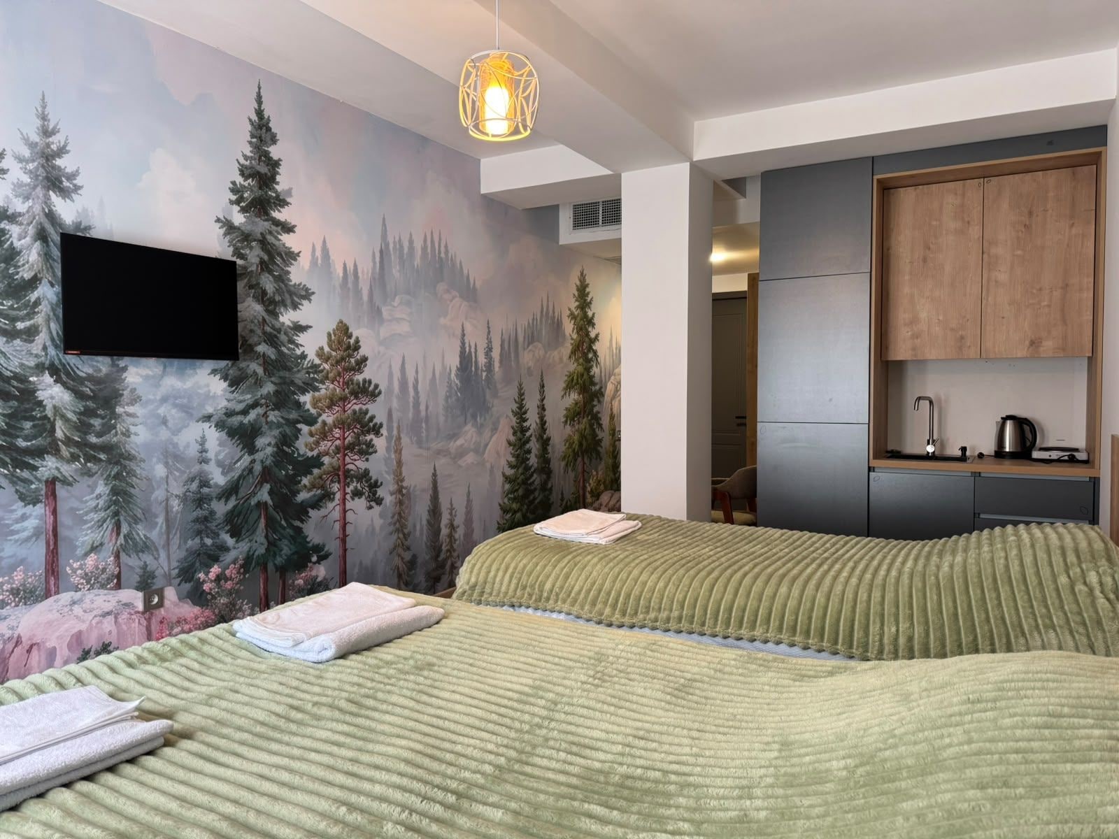 Quarto de hotel com duas camas, televisão fixada na parede, área de cozinha com pia e armários, e um mural com tema de floresta na parede.