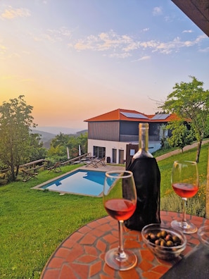 Vista da piscina com uma casa, uma garrafa de vinho e copos sobre uma mesa.
