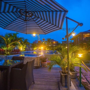 Uma área na beira da piscina com espreguiçadeira, guarda-sol listrado e piscina bem iluminada.