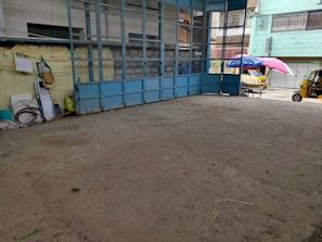 Área externa cercada com chão de terra, um tuk-tuk amarelo e um edifício azul ao fundo.