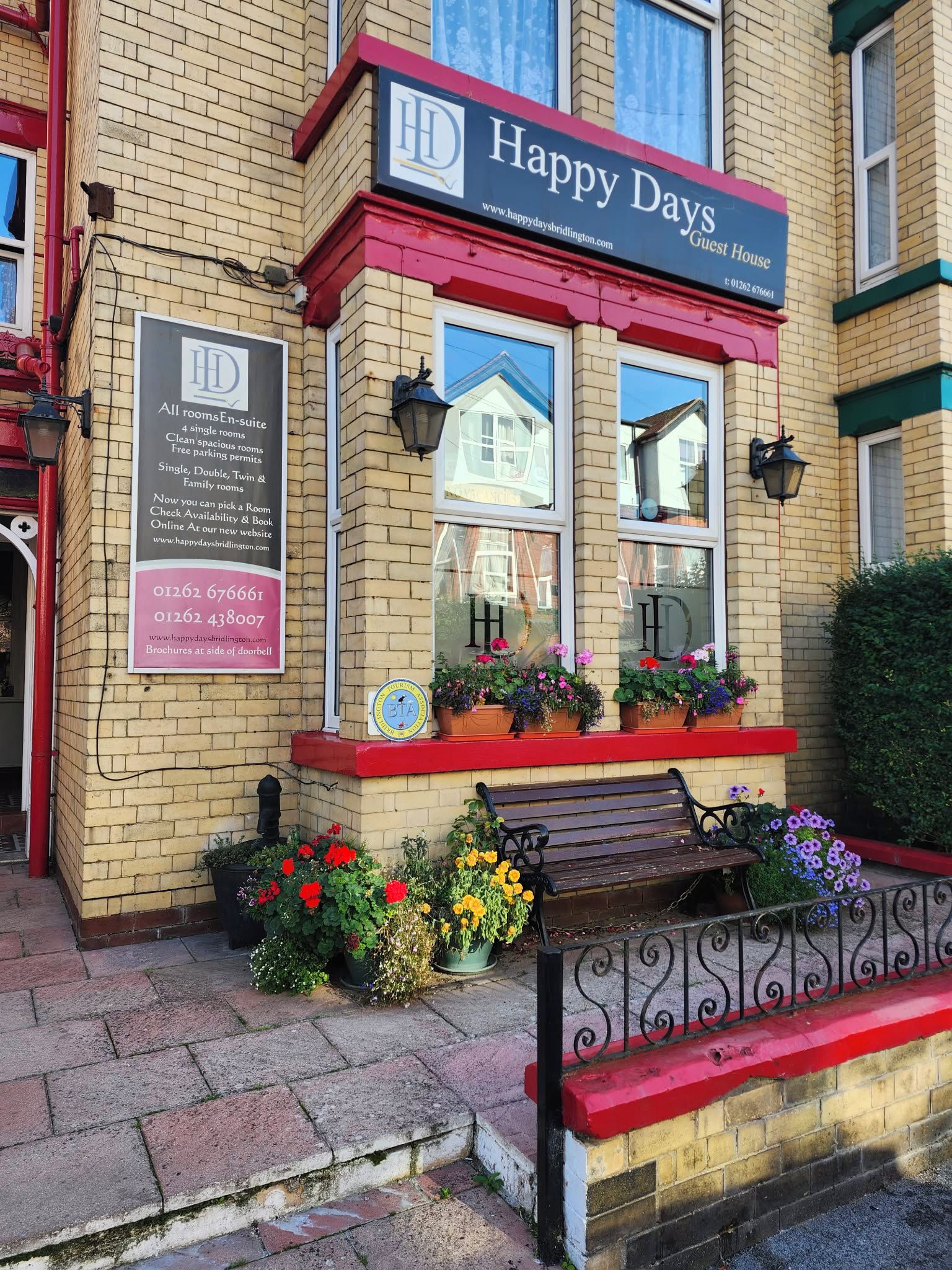 Edifício de tijolos com toldo vermelho e uma placa que diz 'Happy Days Guest House'. Há vasos de flores na calçada e um banco em frente.