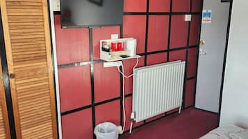 Quarto de hotel com painel de parede em design vermelho e preto, televisão fixada na parede, radiador, lixeira e cama com manta listrada.