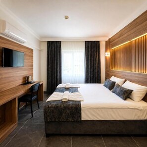 Quarto de hotel com cama, escrivaninha, televisão e luminária fixada na parede.