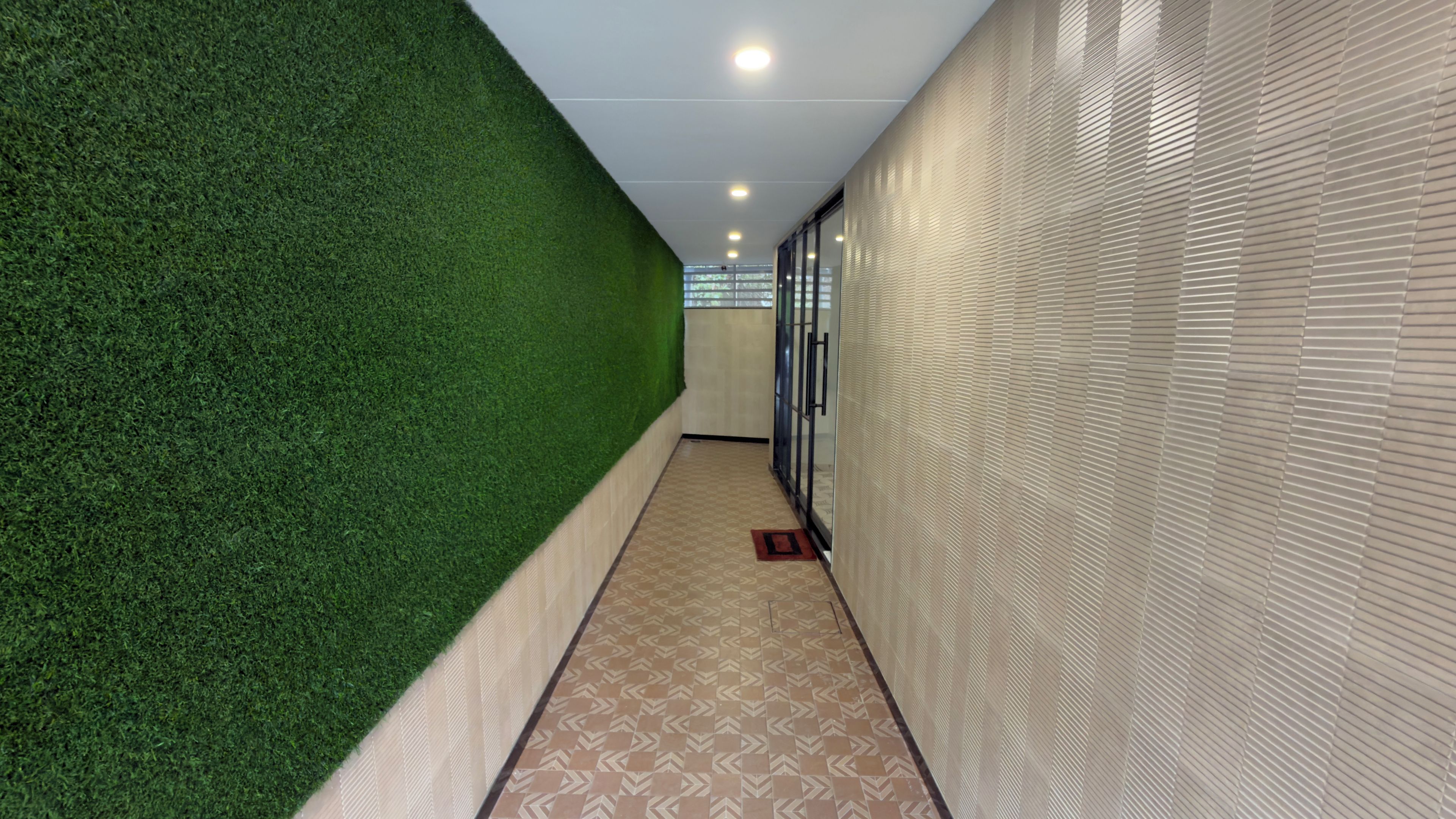 Corredor com parede verde artificial, piso estampado e porta com painel de vidro.