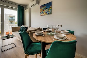 Uma sala de jantar moderna com mesa redonda de madeira, cadeiras estofadas verdes e vista para uma varanda com paisagem urbana.