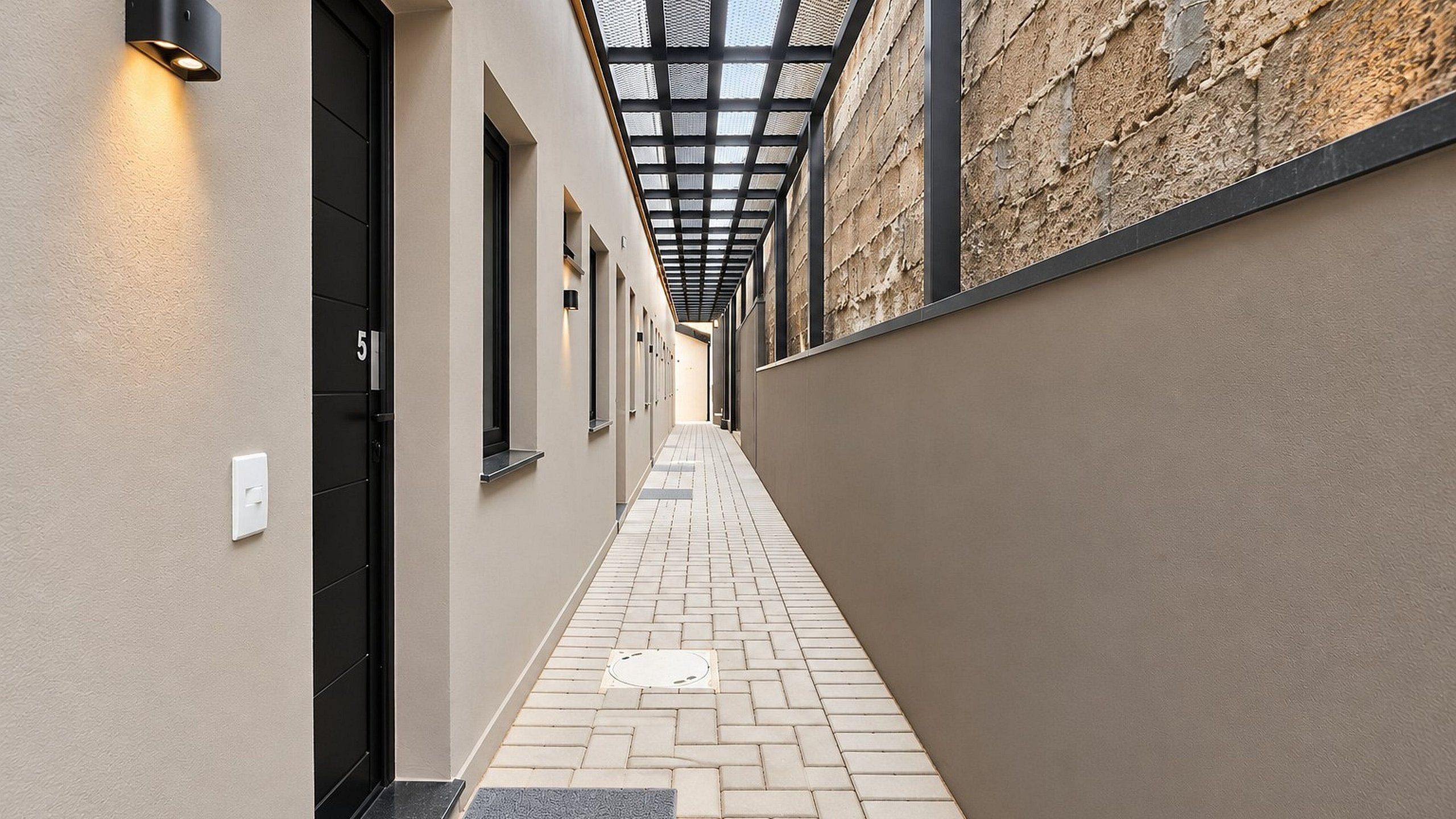 Um corredor com piso de azulejos, uma porta preta com número e um luminário fixado na parede.