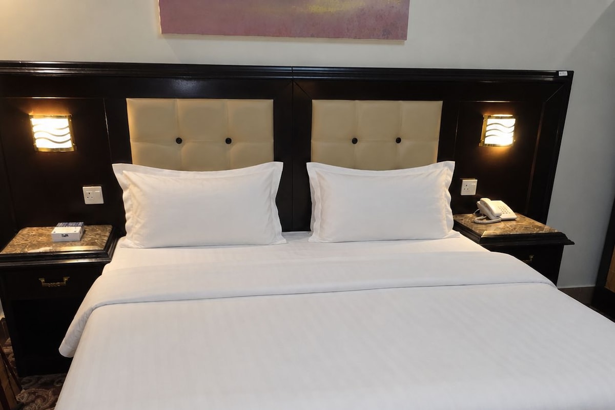 Quarto de hotel com uma cama grande, duas mesas de cabeceira, um telefone fixado na parede e um quadro sobre a cama.