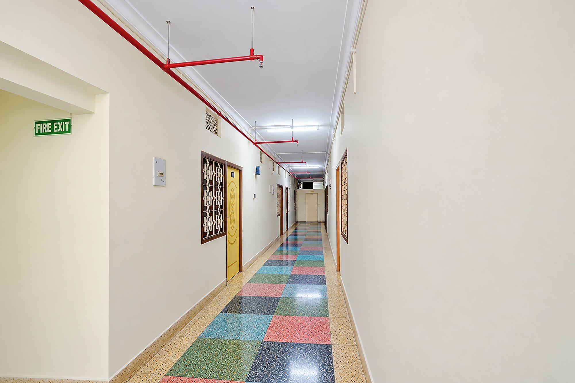 Um corredor com sinal de saída de incêndio, uma porta com grade decorativa e um chão com ladrilhos coloridos.