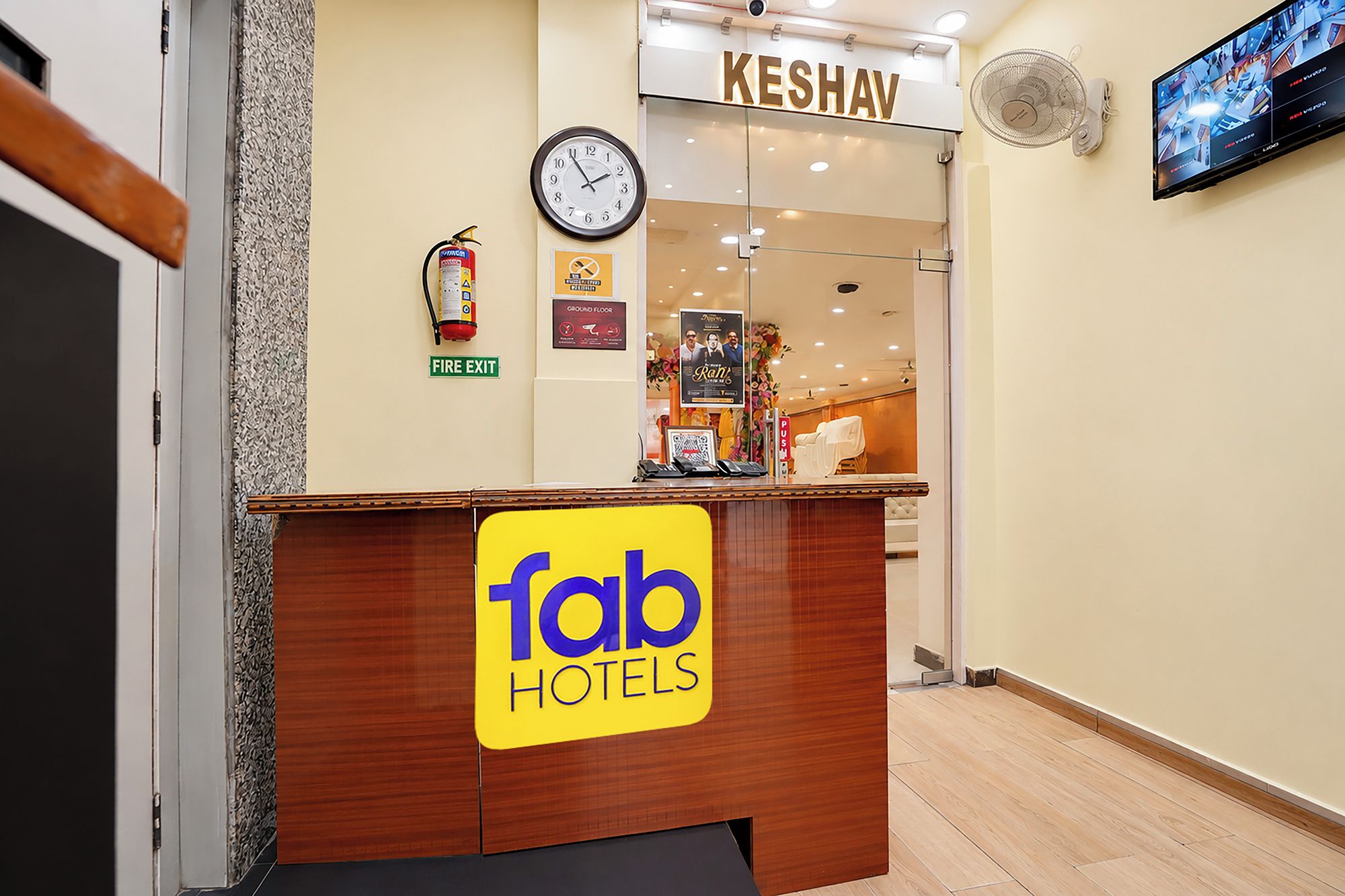 Bancada de recepção de hotel com um relógio, placa de saída de incêndio e sinalização para 'Keshav Hotels'.