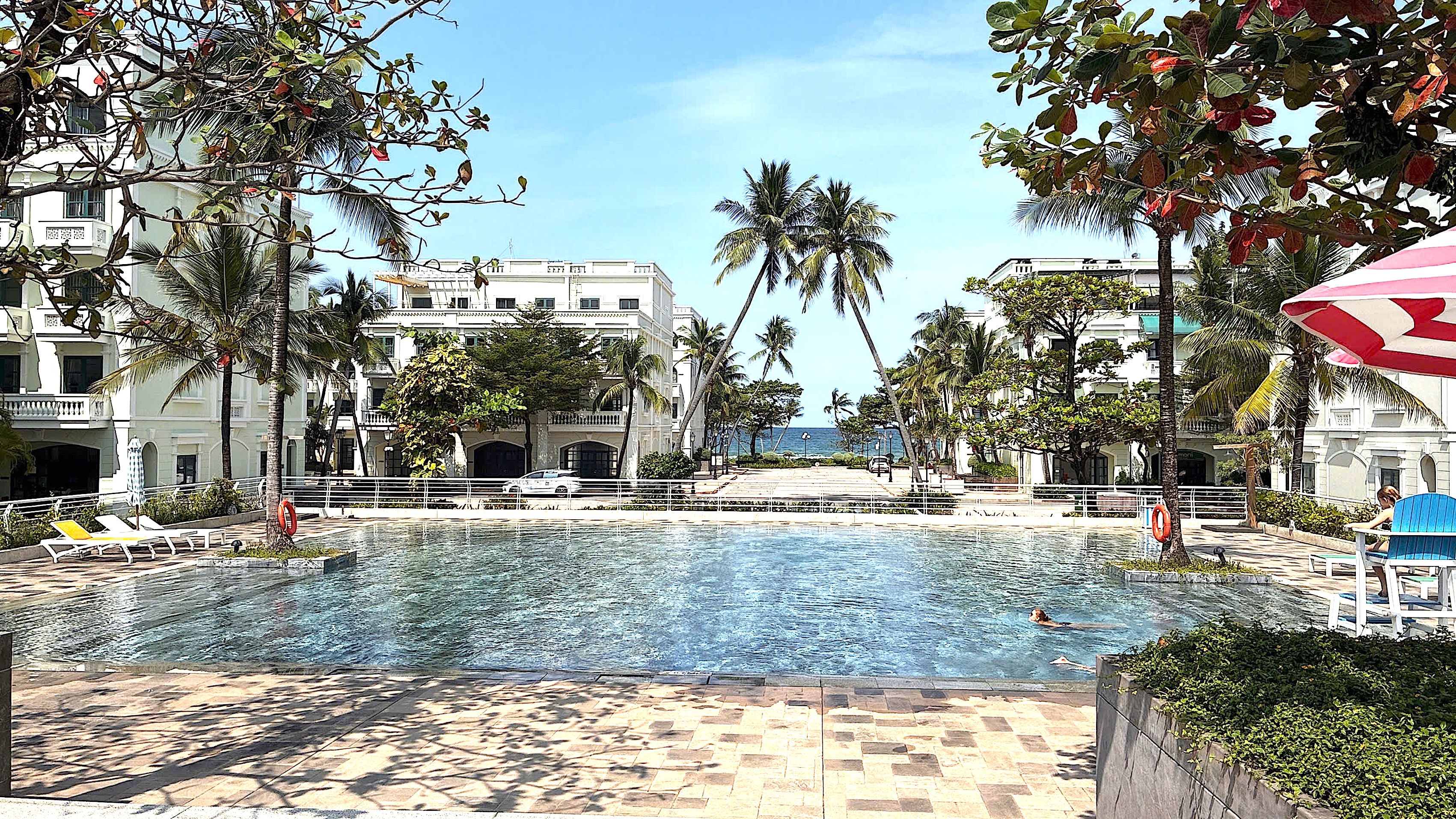 Piscina com espreguiçadeiras e palmeiras, cercada por edifícios.