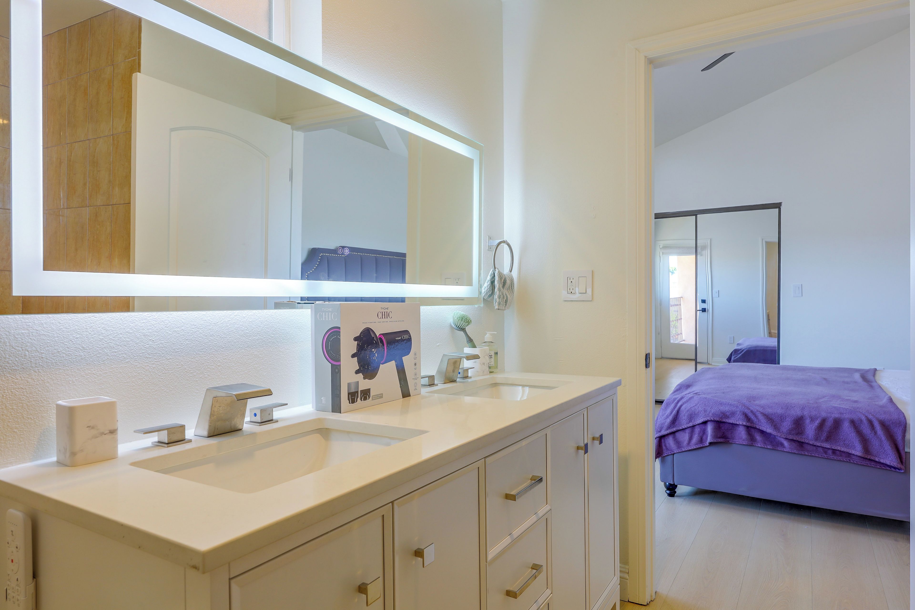 Um banheiro com um espelho grande, duas pias, um porta-toalhas e, no quarto vizinho, uma cama com um cobertor roxo.