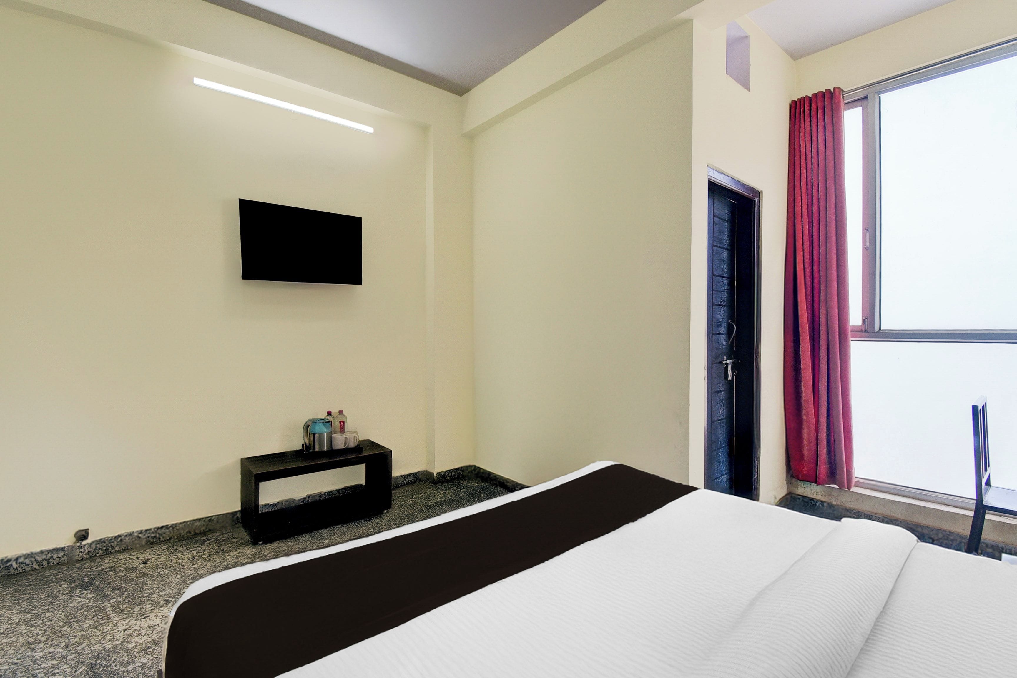 Quarto de hotel com cama, uma mesinha com chaleira e xícara, TV de tela plana na parede e uma janela com cortinas.