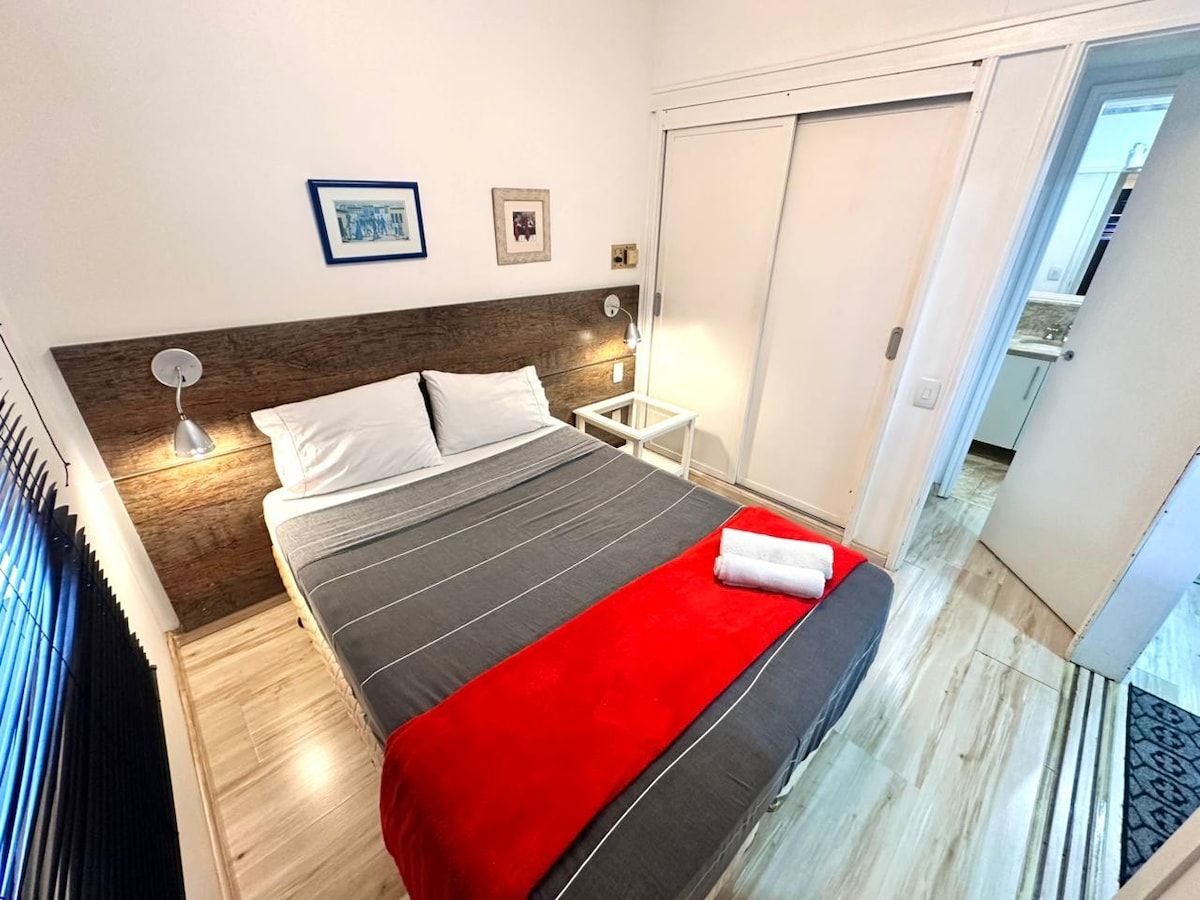 Cama bem arrumada com manta vermelha e travesseiros brancos, abajur e cabeceira de madeira.