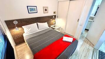 Cama bem arrumada com manta vermelha e travesseiros brancos, abajur e cabeceira de madeira.