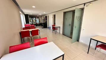 Uma área de refeições com mesas e cadeiras, uma cozinha ao fundo e uma porta que dá para outro cômodo.