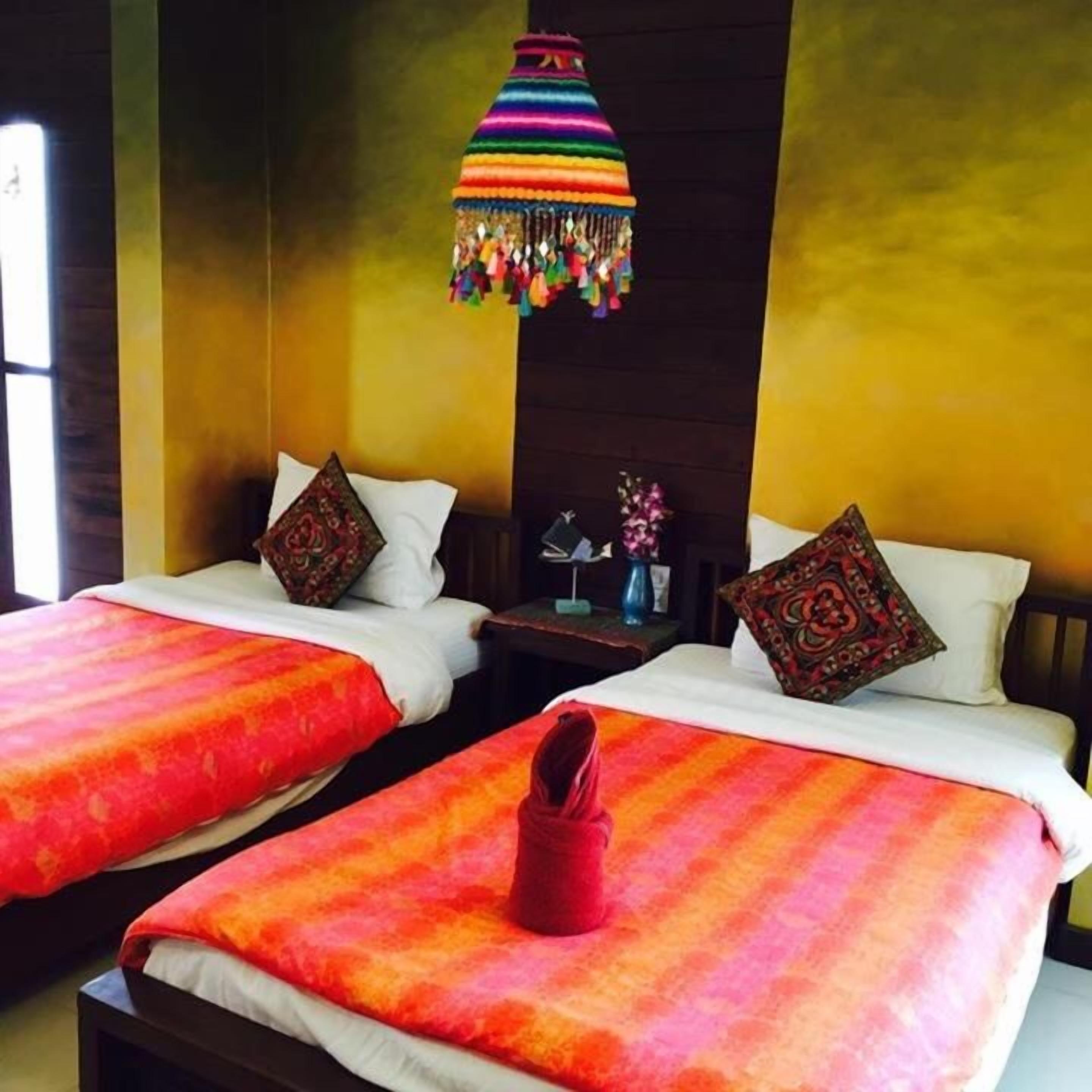 Quarto de hotel com duas camas, cada uma com colcha listrada em vermelho e laranja, uma luminária colorida pendente e uma mesa pequena com um vaso de flores.