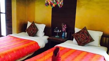 Quarto de hotel com duas camas, cada uma com colcha listrada em vermelho e laranja, uma luminária colorida pendente e uma mesa pequena com um vaso de flores.