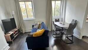 Uma sala de estar moderna com piso de madeira escura, um sofá azul com almofadas amarelas, uma mesa de jantar com cadeiras e uma TV de tela plana.