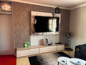 Uma sala de estar moderna com televisão de tela plana presa a uma parede de mármore, um móvel de entretenimento de madeira abaixo, um tapete preto e branco e uma mala preta.