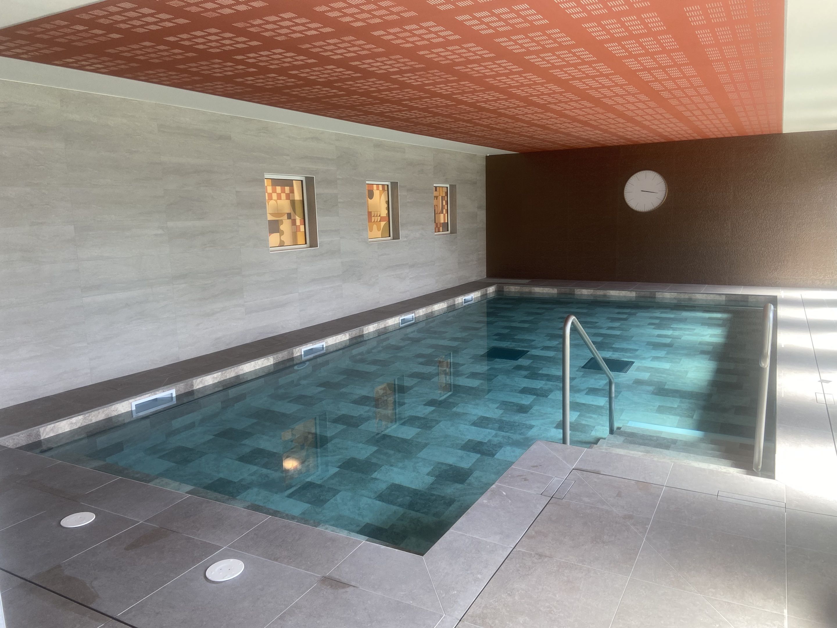 Piscina interna com chão xadrez, guarda-corpo de metal e relógio na parede.