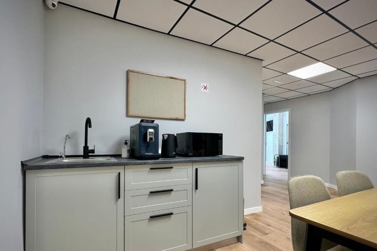 Uma cozinha compacta com pia, armários e cafeteira. Há um quadro na parede e uma porta com placa vermelha em formato de 'X'.