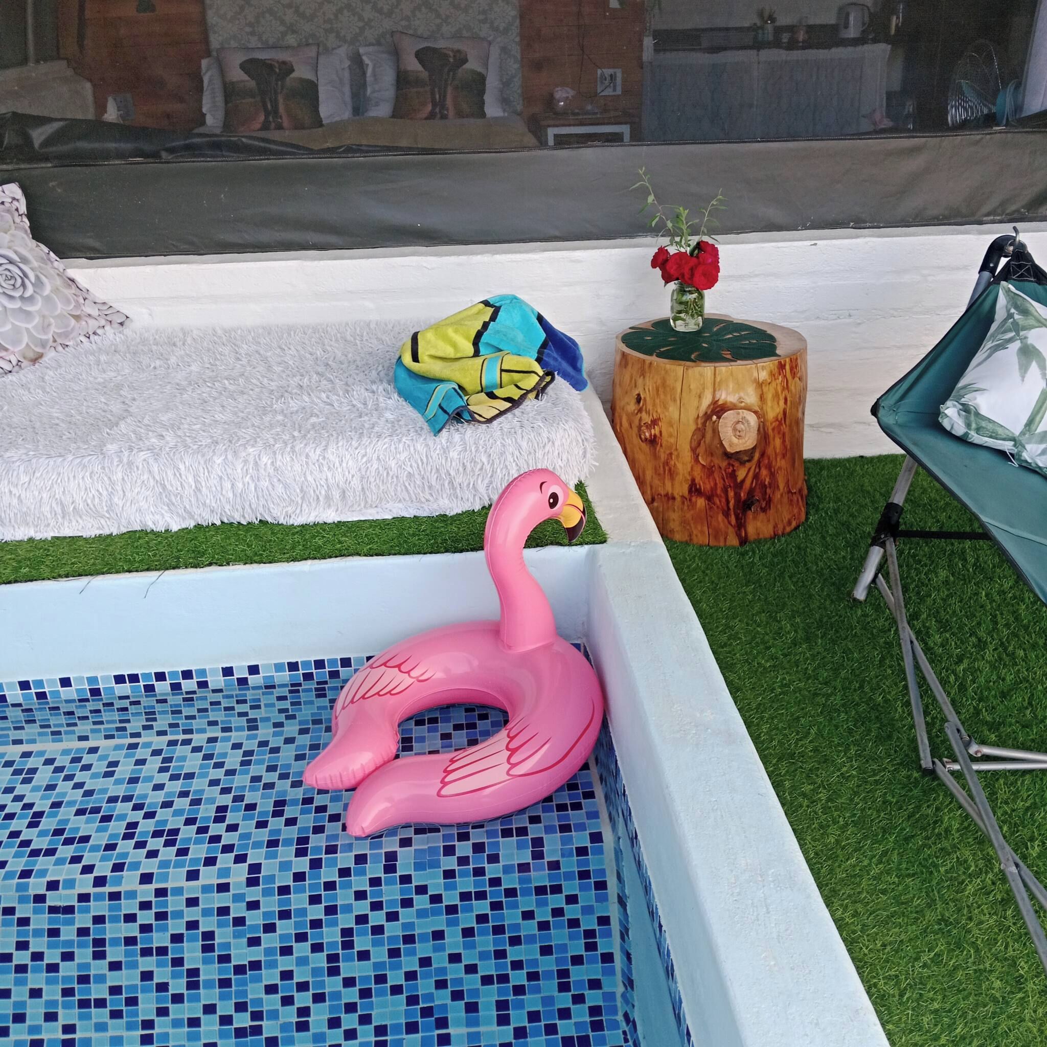Uma piscina com um flutuador em formato de flamingo rosa, um toco de madeira com um vaso de flores e uma cadeira dobrável verde.