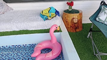 Uma piscina com um flutuador em formato de flamingo rosa, um toco de madeira com um vaso de flores e uma cadeira dobrável verde.