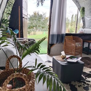 Tenda com vista para um gramado verde e árvores. No interior, um cesto de vime, uma cadeira trançada e uma mesa com uma revista.