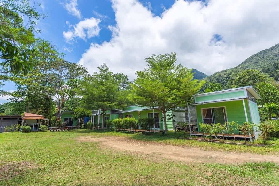 Fileira de cabanas verdes com um caminho de terra em frente, cercada por árvores e uma montanha ao fundo.