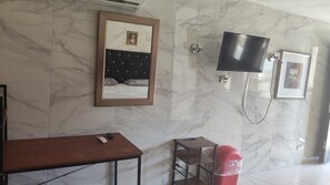 Quarto de hotel com parede de mármore, quadro emoldurado, TV de tela plana fixada na parede, mesa de madeira pequena e lixeira.