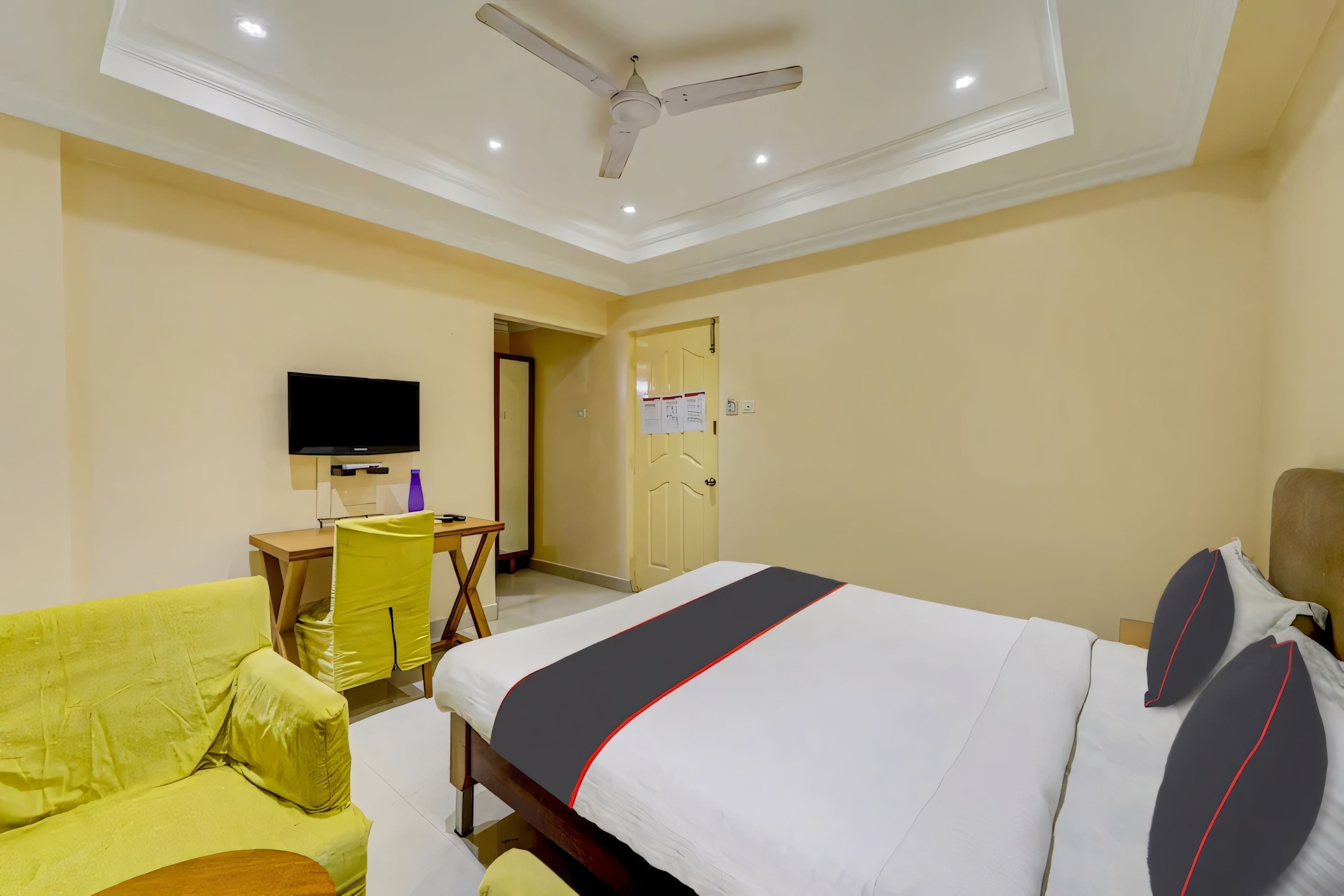 Quarto de hotel com cama, escrivaninha, cadeira, televisão e ventilador de teto.