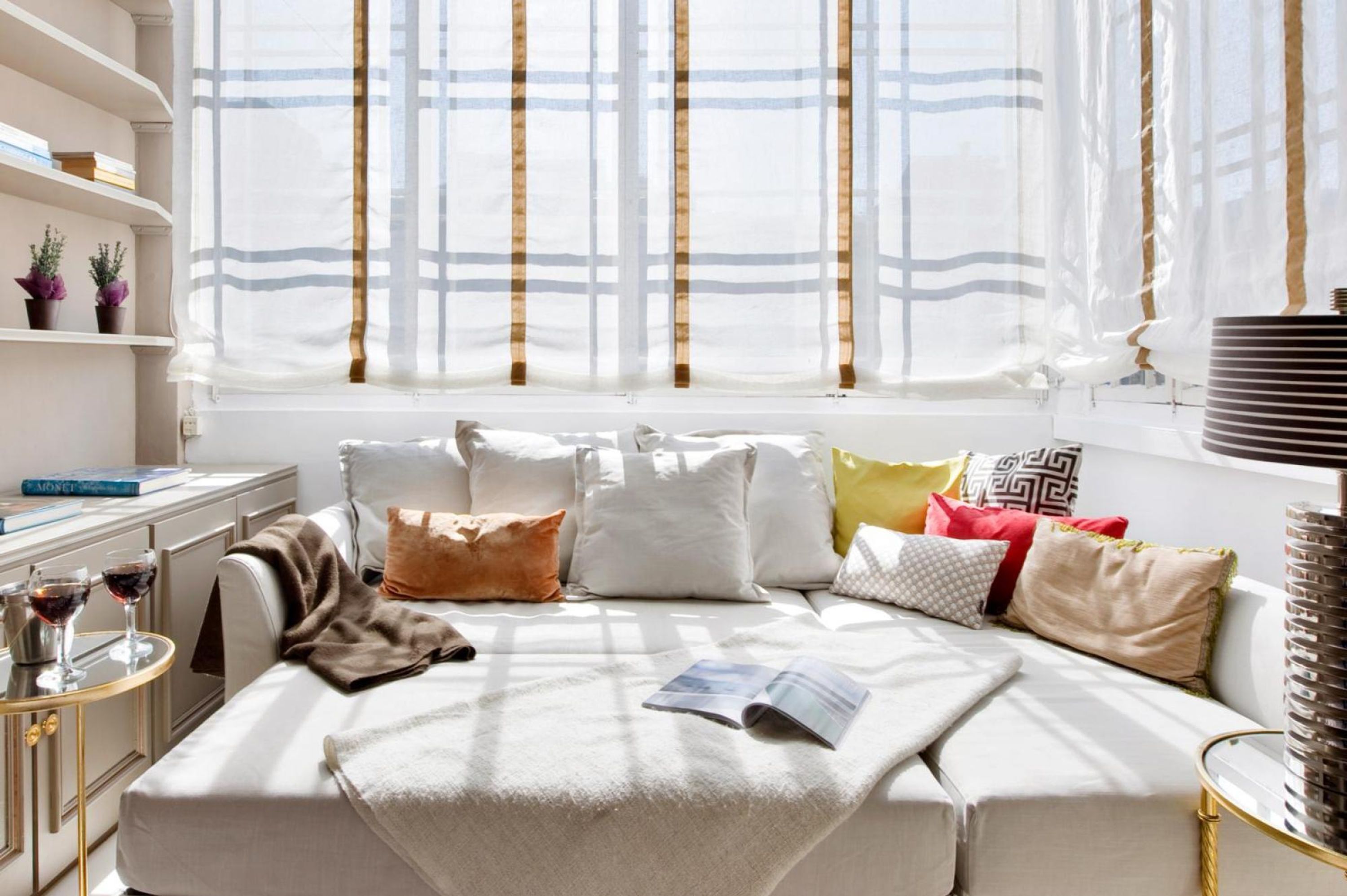 Quarto bem iluminado com cama, almofadas, uma revista, uma mesinha com copos e uma janela com cortinas finas.