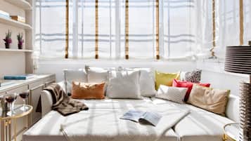 Quarto bem iluminado com cama, almofadas, uma revista, uma mesinha com copos e uma janela com cortinas finas.