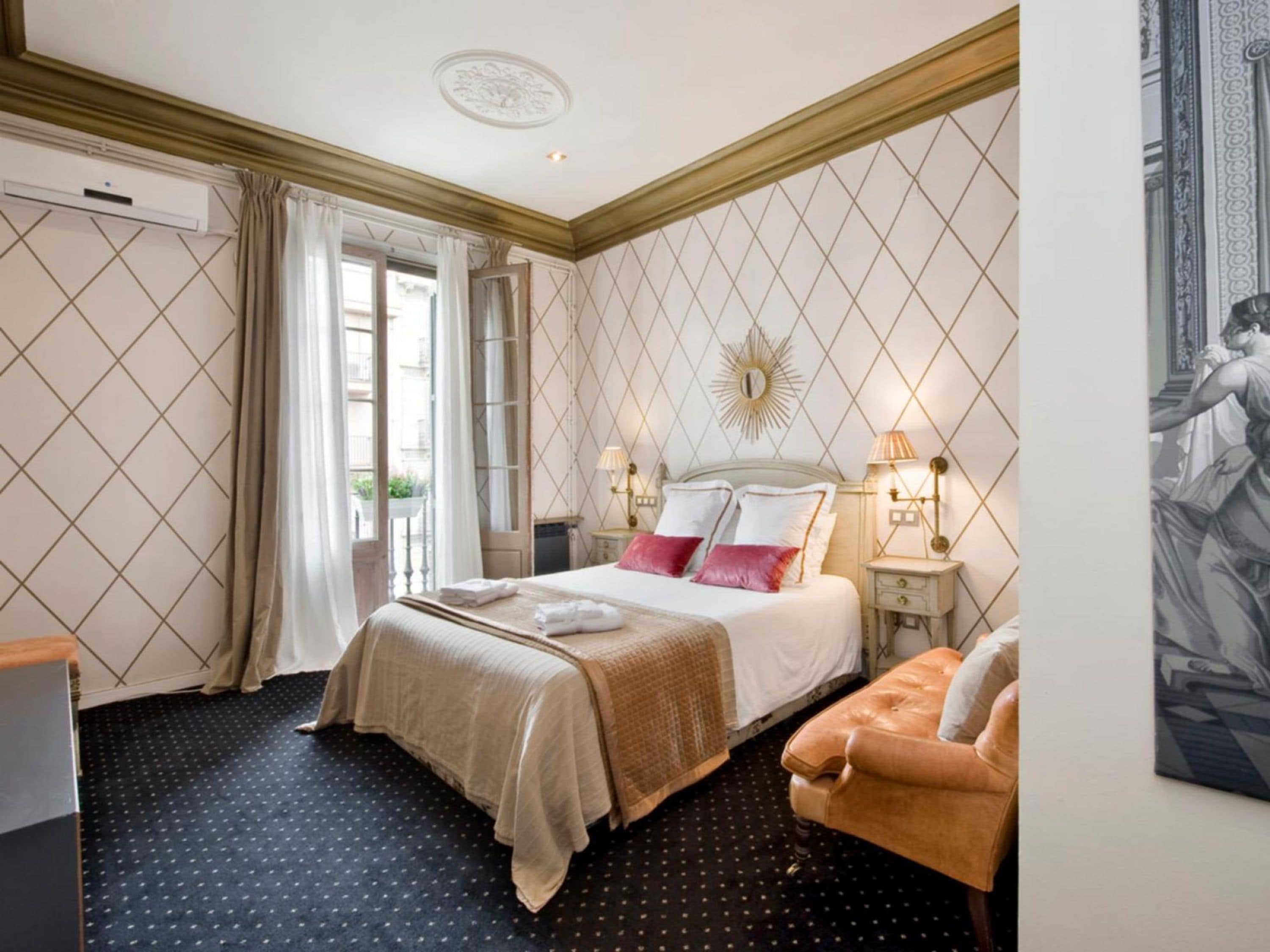 Quarto de hotel com uma cama, uma cadeira, uma mesinha de cabeceira e uma luminária na parede.