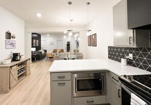 Cozinha moderna com ilha central, forno embutido e micro-ondas. Há uma área de refeições com mesa e cadeiras, e um cantinho do café com um letreiro.