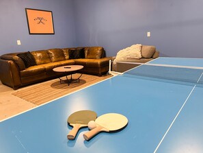 Um quarto com uma mesa de pingue-pongue azul, duas raquetes de pingue-pongue, um sofá de couro marrom, uma pequena mesa redonda e um quadro na parede.