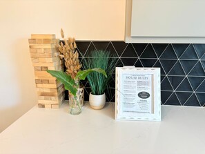 Placa com as regras da casa, texto e senha de WiFi, emoldurada e colocada sobre uma bancada branca, ao lado de um vaso com plantas e um painel decorativo.