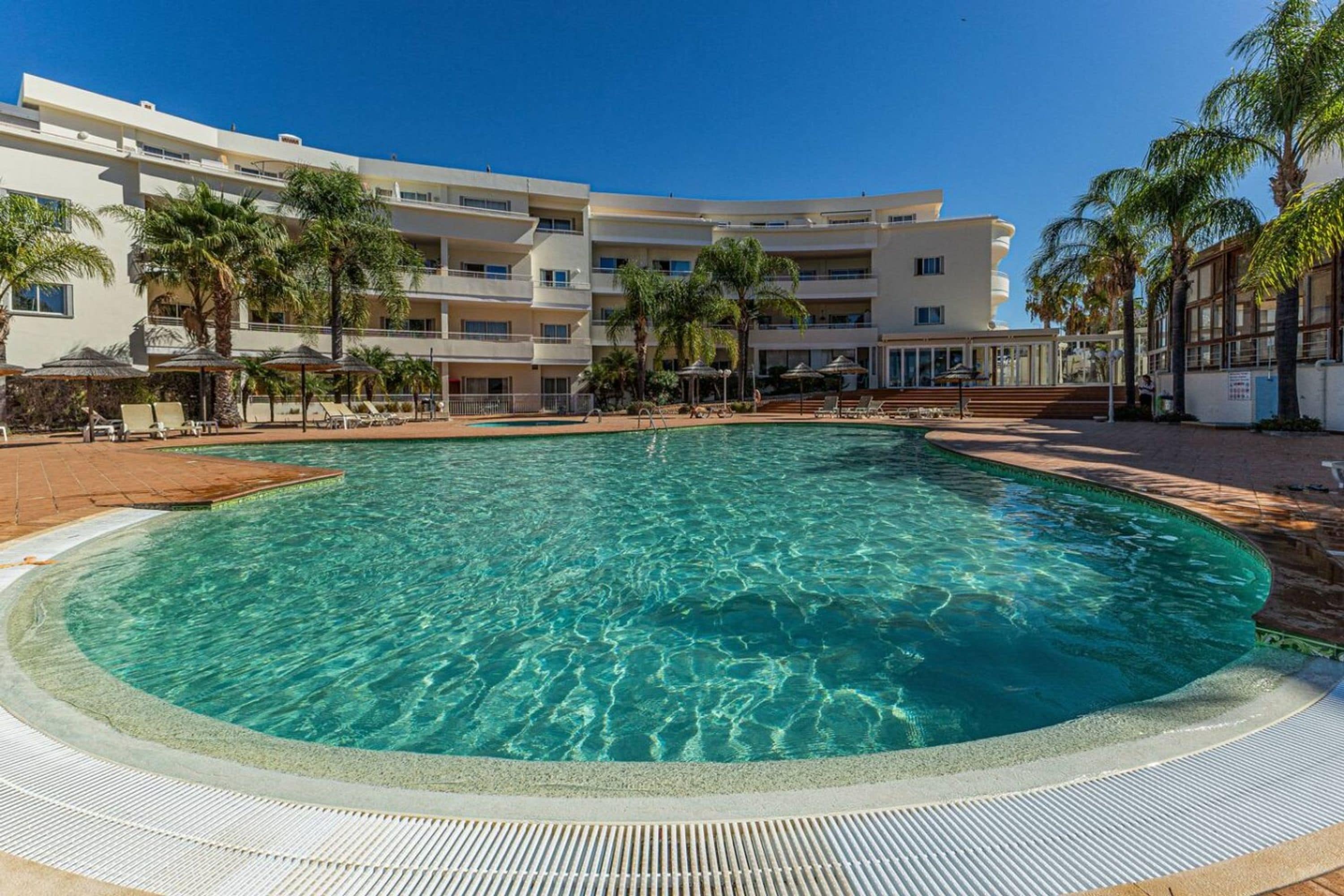 Piscina com águas cristalinas, rodeada por espreguiçadeiras e guarda-sóis, com um edifício de vários andares ao fundo.