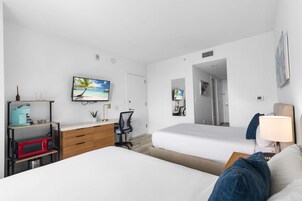 Quarto de hotel com duas camas, uma escrivaninha com cadeira, máquina de café, televisão e banheiro visível por uma porta aberta.