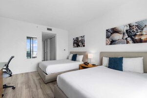 Quarto de hotel com duas camas, uma escrivaninha, uma cadeira, um abajur e obras de arte nas paredes.