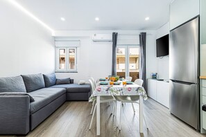 Uma sala de estar moderna com um sofá seccional cinza, uma mesa de jantar com cadeiras, uma área de cozinha com geladeira e uma TV de tela plana na parede.