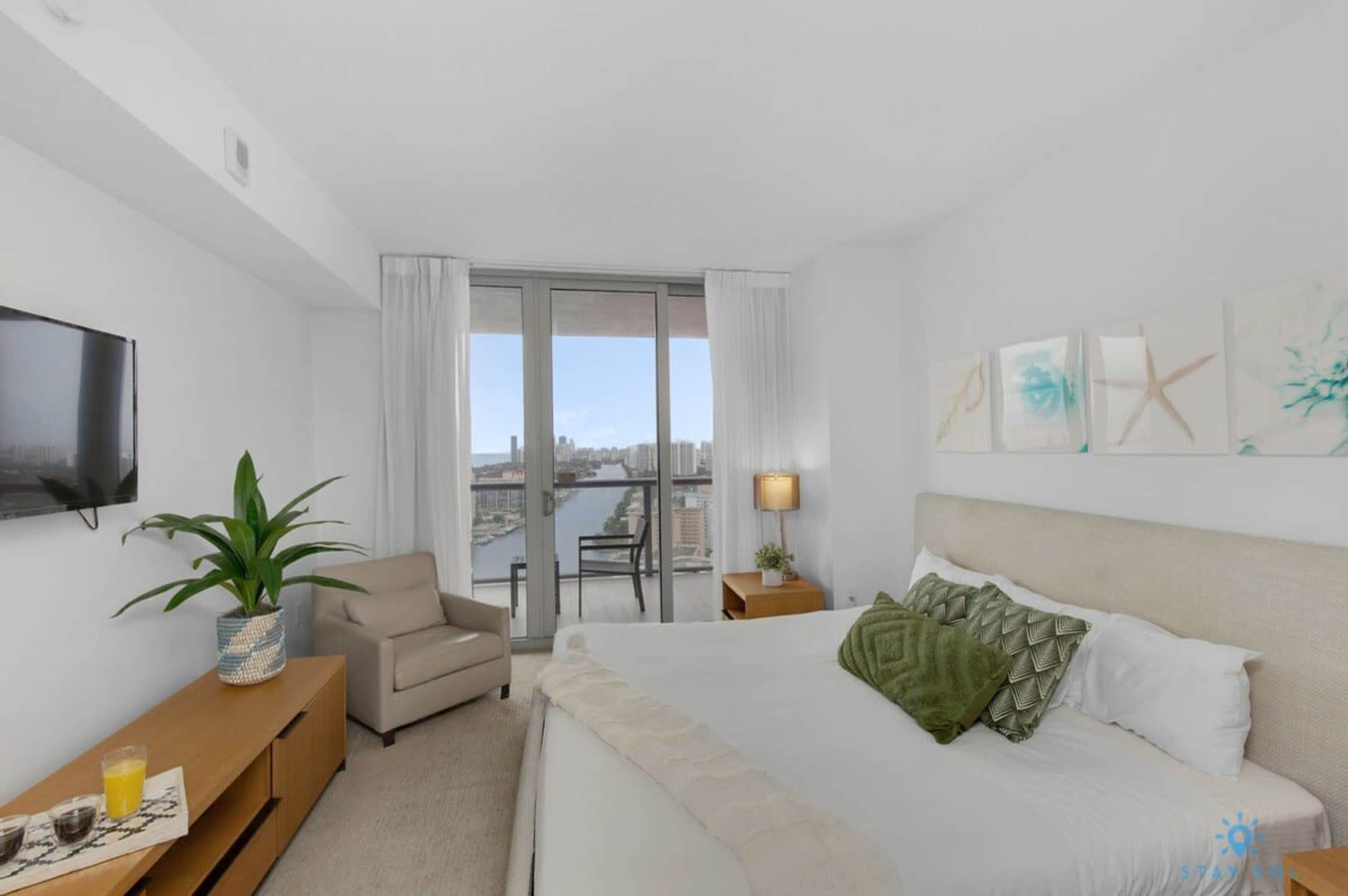 Quarto de hotel com cama, televisão, planta, cadeira, mesa de cabeceira e obras de arte na parede.