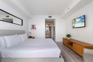 Quarto de hotel moderno com uma cama grande, um console de madeira, uma TV e um banheiro visível por uma porta aberta.