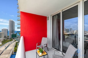 Varanda com parede vermelha, duas cadeiras, uma mesa pequena com uma planta e vista para a cidade.